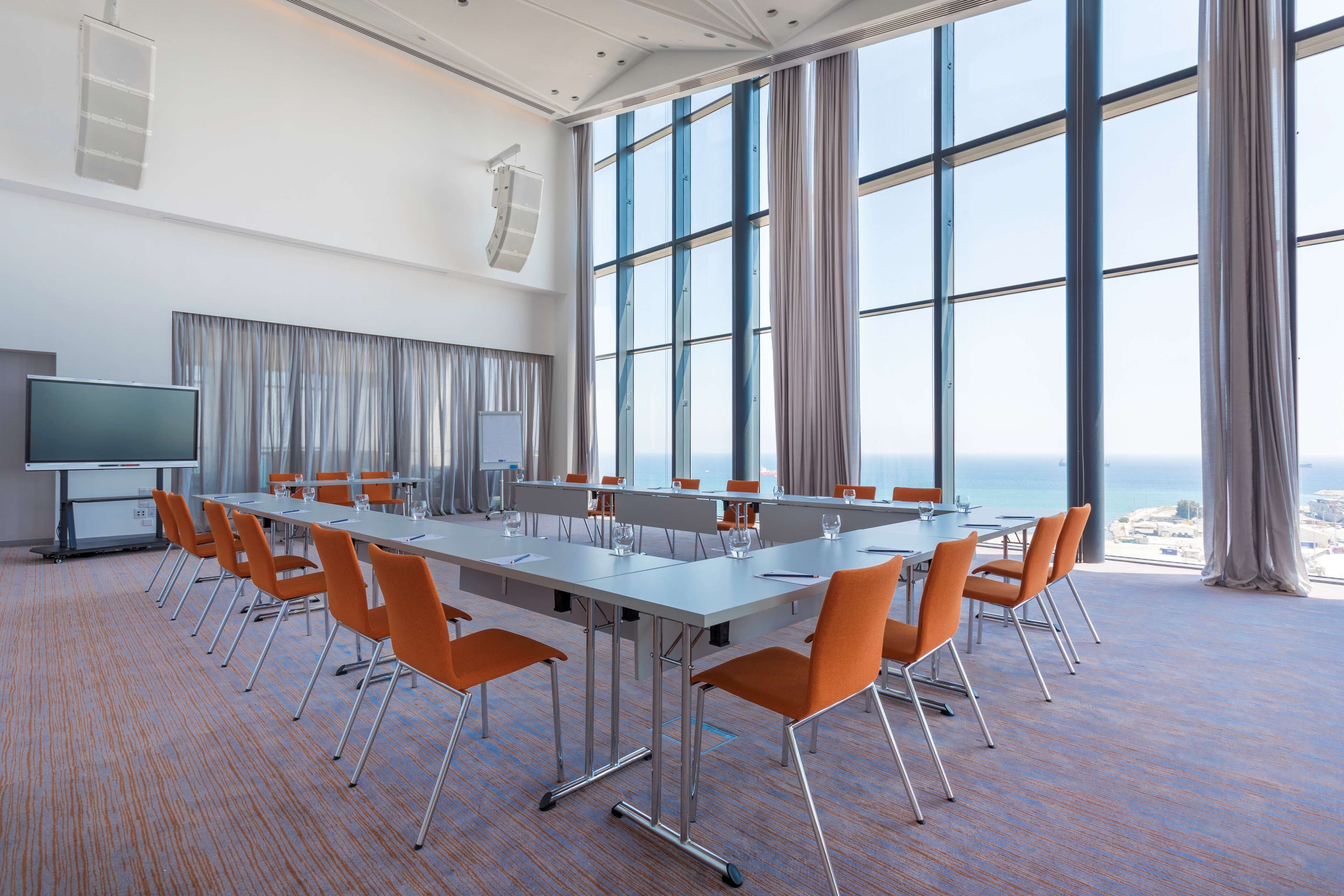 Uma sala de conferências com uma mesa comprida, cadeiras laranja, tela de projetor e grandes janelas com vista para o mar.