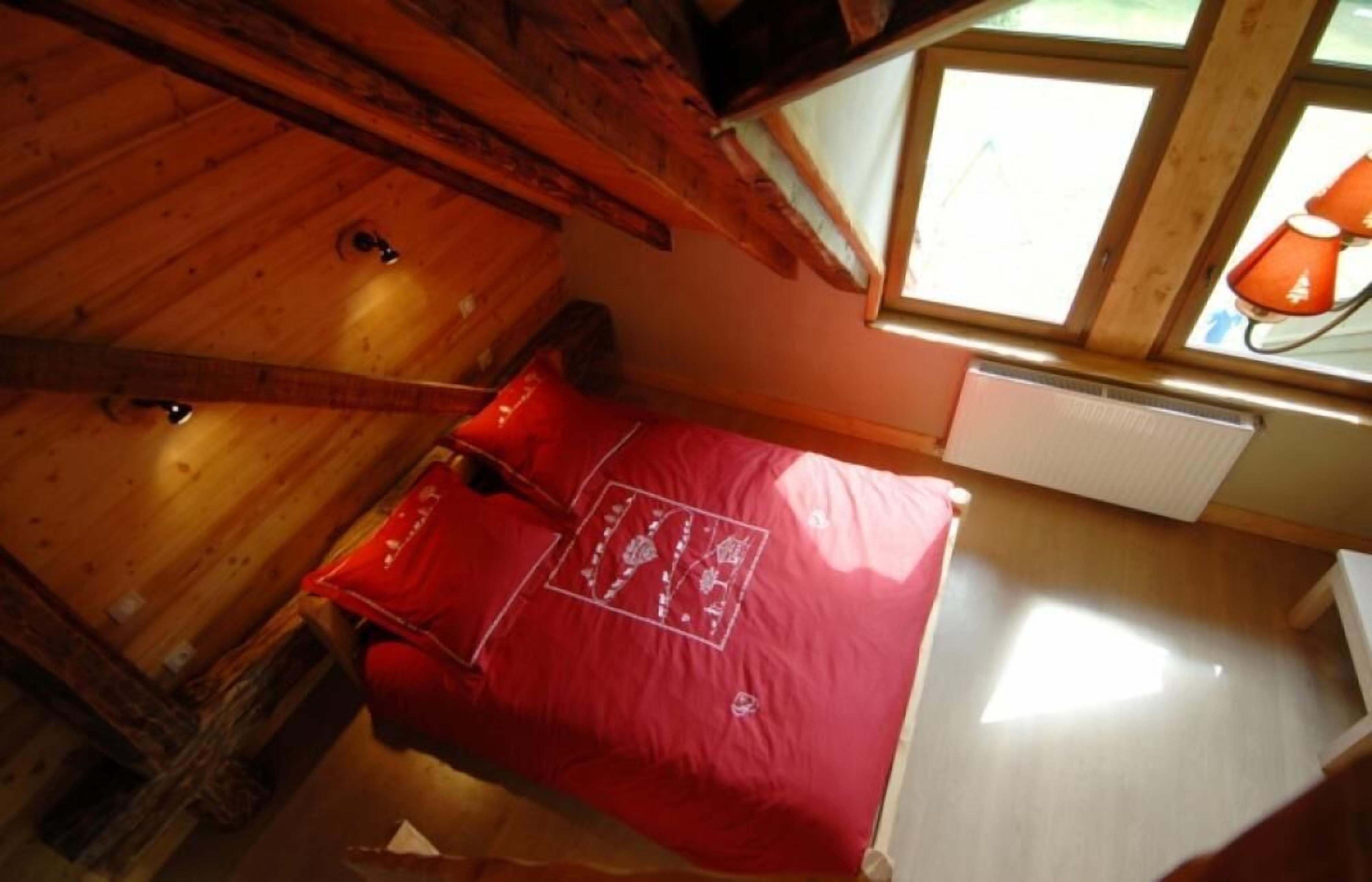 Um quarto pequeno e acolhedor com teto de madeira, colcha vermelha e janela com vista para o verde lá fora.