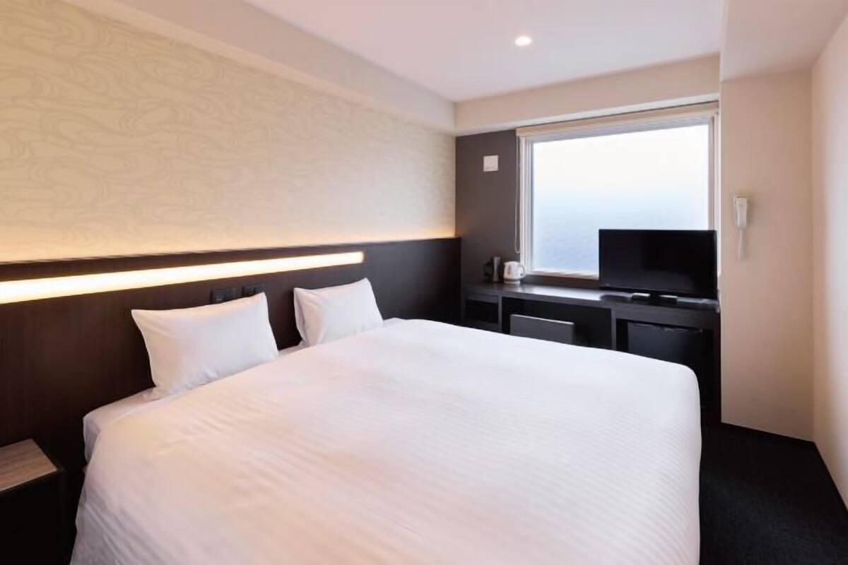 Quarto de hotel com uma cama grande, uma escrivaninha e uma televisão, além de uma janela.