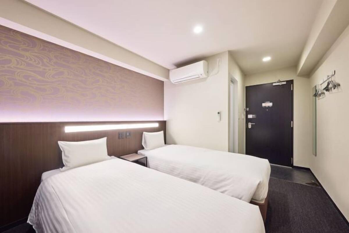 Quarto de hotel com duas camas, ar condicionado fixado na parede, porta e uma pequena mesa de cabeceira.
