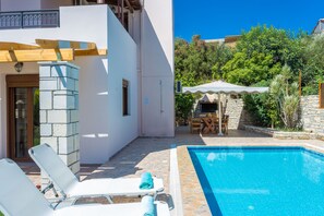 Uma área de piscina com espreguiçadeiras e uma mesa, cercada por paredes de pedra e vegetação.