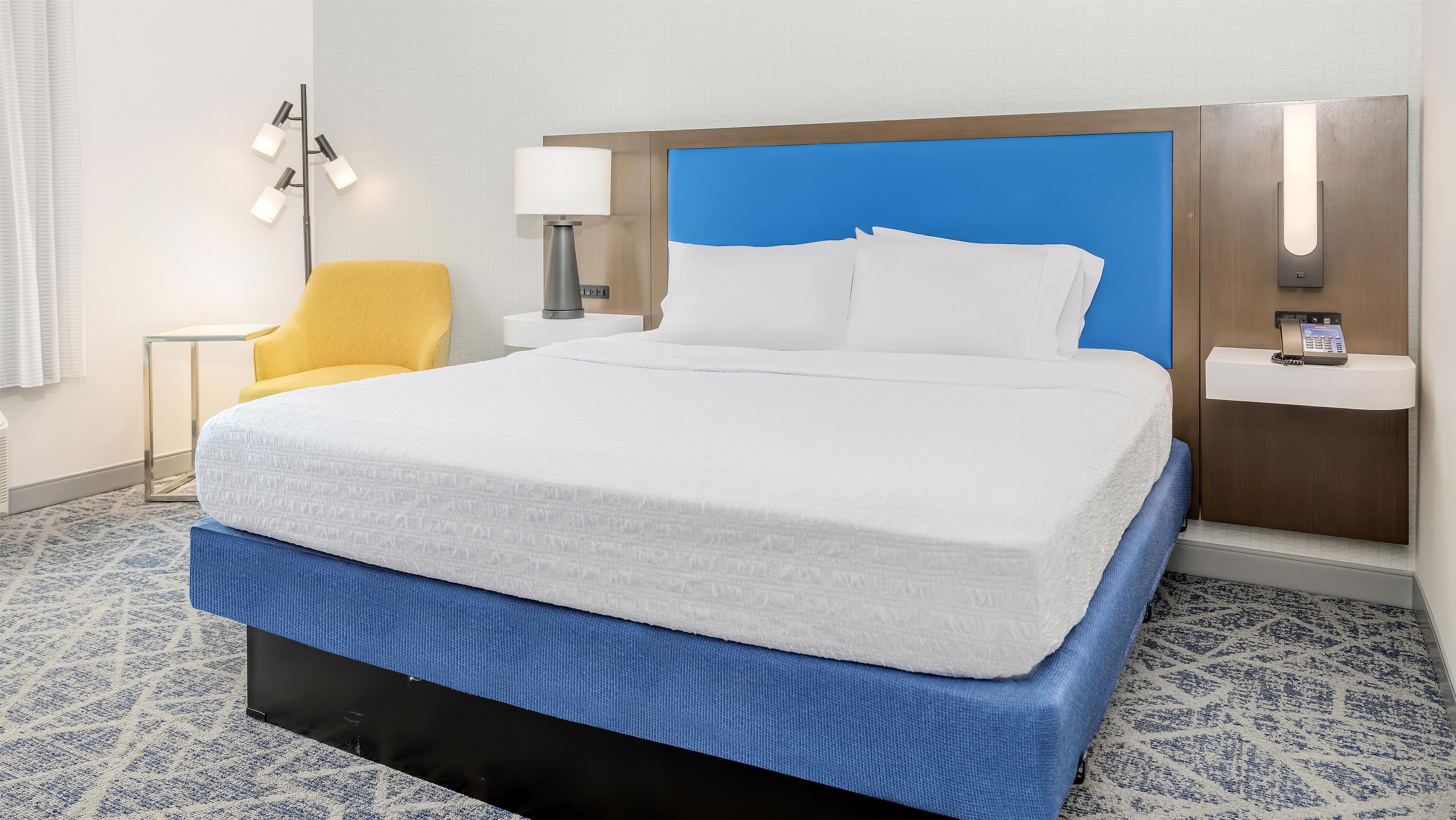Quarto de hotel com cama grande, mesa de cabeceira com luminária, cadeira e telefone fixado na parede.