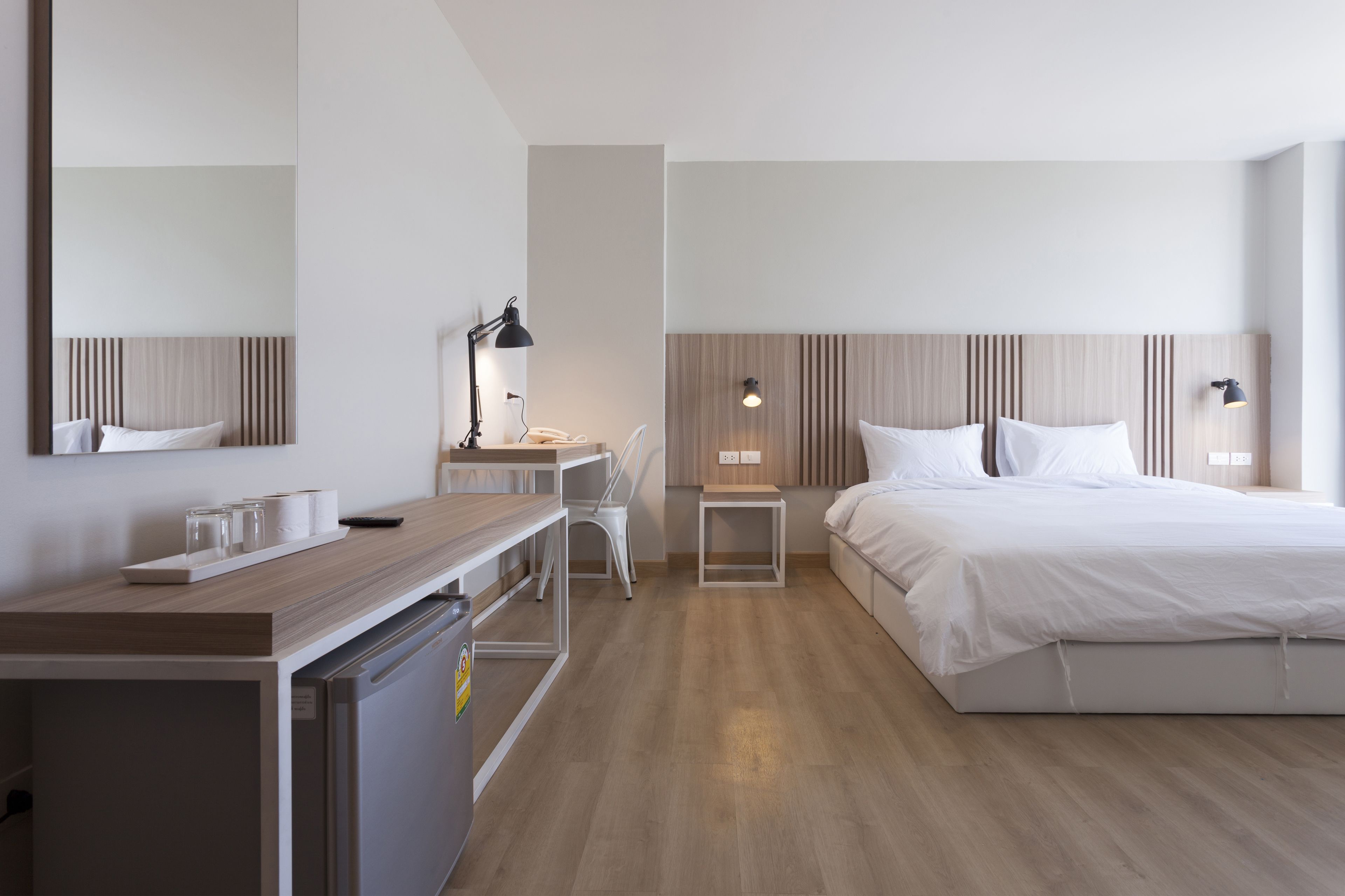 Quarto de hotel moderno com piso de madeira, uma cama, uma escrivaninha com cadeira, uma pequena mesa e um frigorífico.
