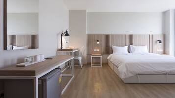 Quarto de hotel moderno com piso de madeira, uma cama, uma escrivaninha com cadeira, uma pequena mesa e um frigorífico.