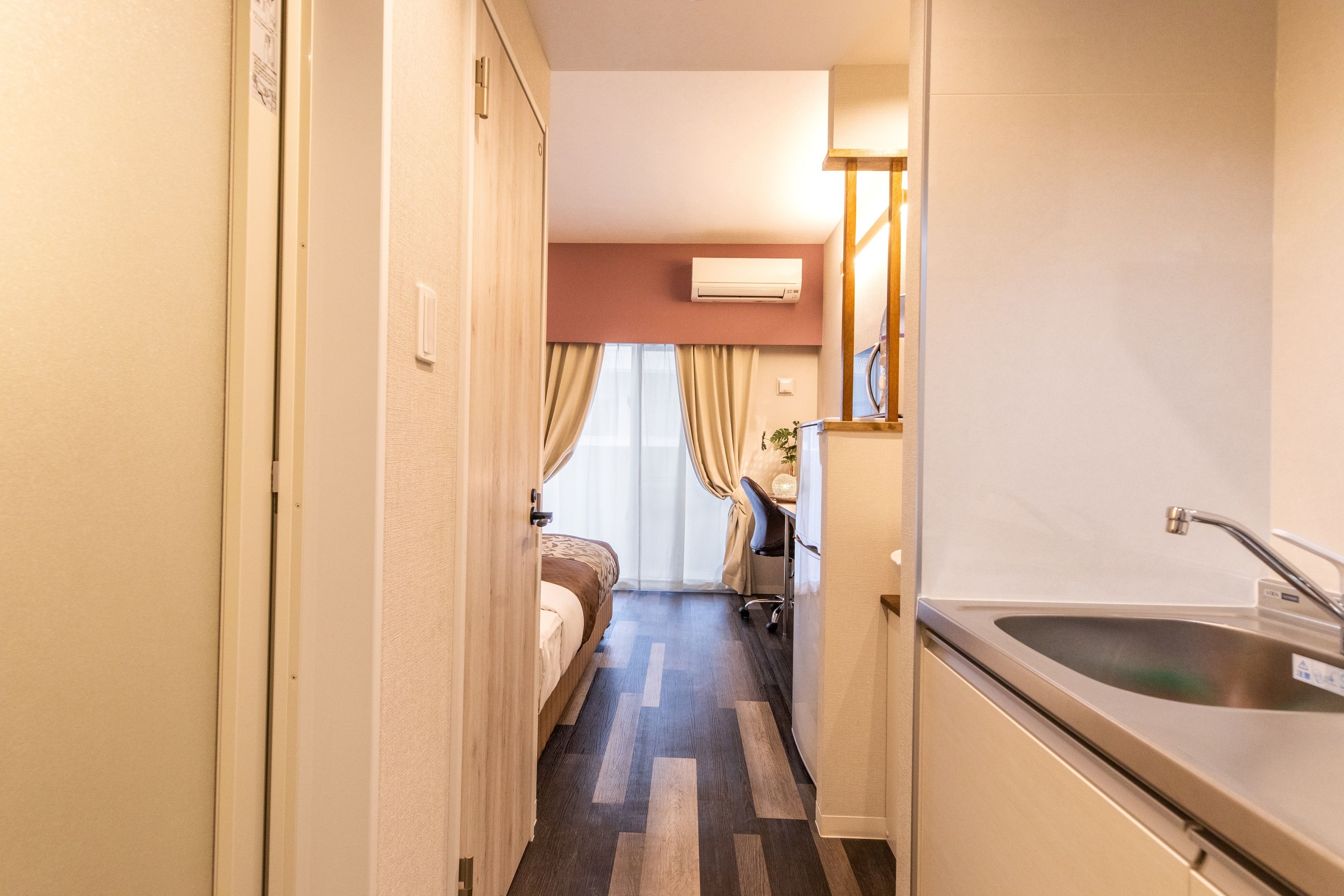 Corredor estreito que leva a um quarto com uma cama, uma escrivaninha e uma pia.
