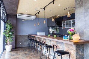 Uma área de bar moderna com um balcão comprido, vários bancos, luminárias pendentes e um vaso com planta.