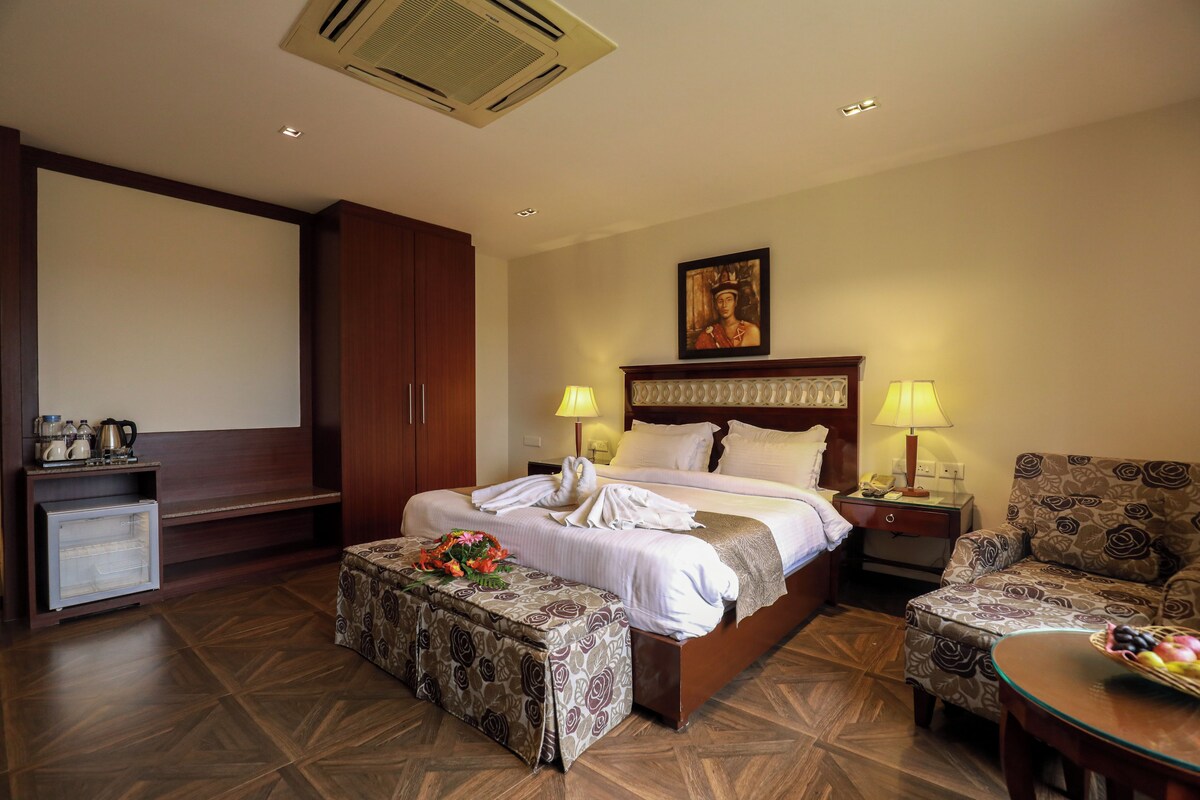 Quarto de hotel com uma cama grande, duas luminárias de cabeceira, um guarda-roupa de madeira, uma pequena mesa com um vaso de flores e uma poltrona estampada.