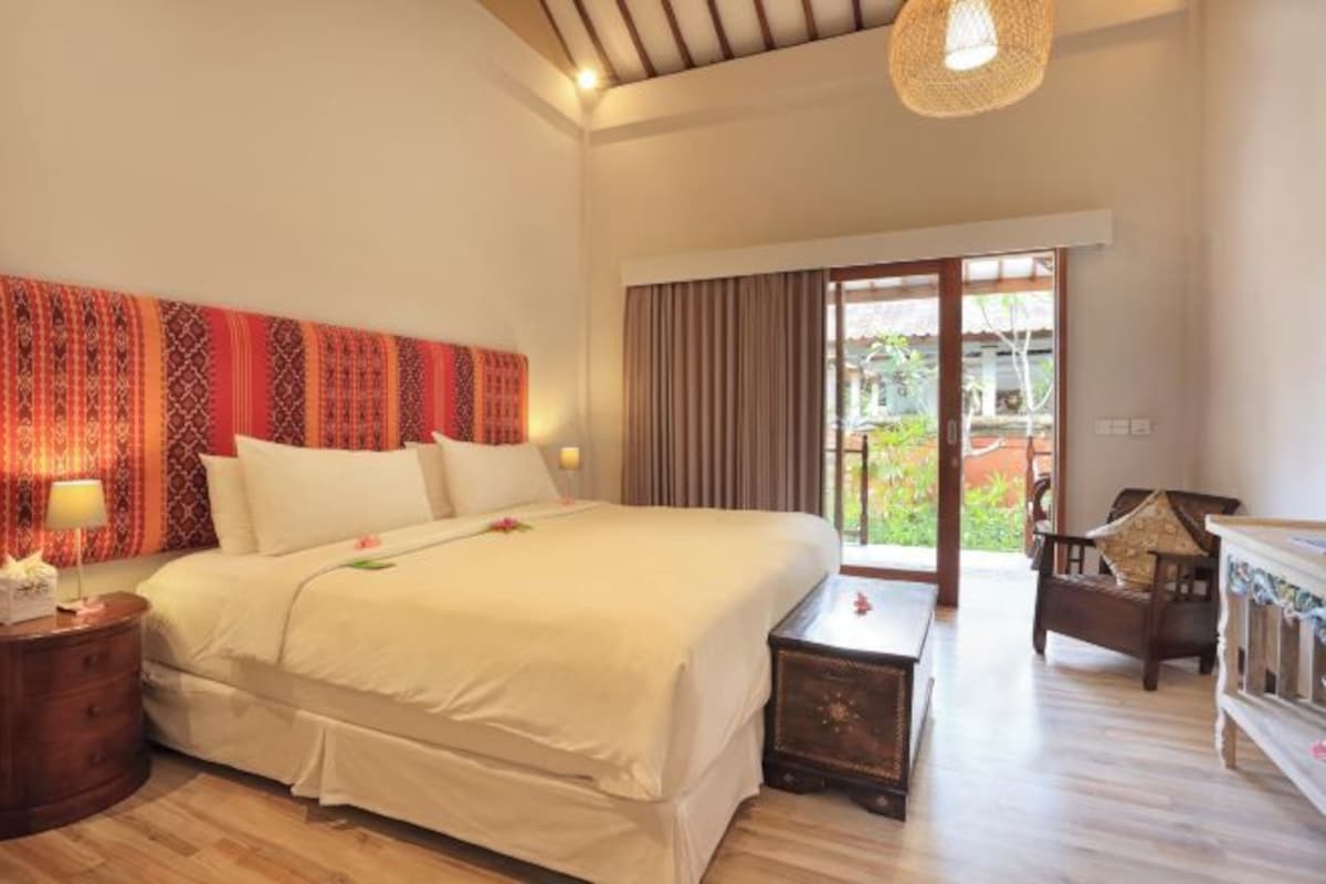 Quarto de hotel com uma cama grande, uma mesinha de cabeceira de madeira, uma pequena mesa, uma cadeira e vista para o exterior através de uma porta deslizante.