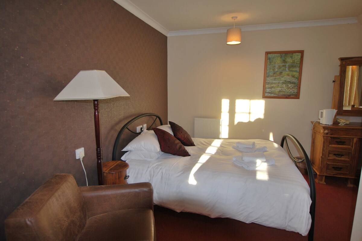Quarto de hotel com cama, uma cadeira, abajur, cômoda e um quadro na parede.
