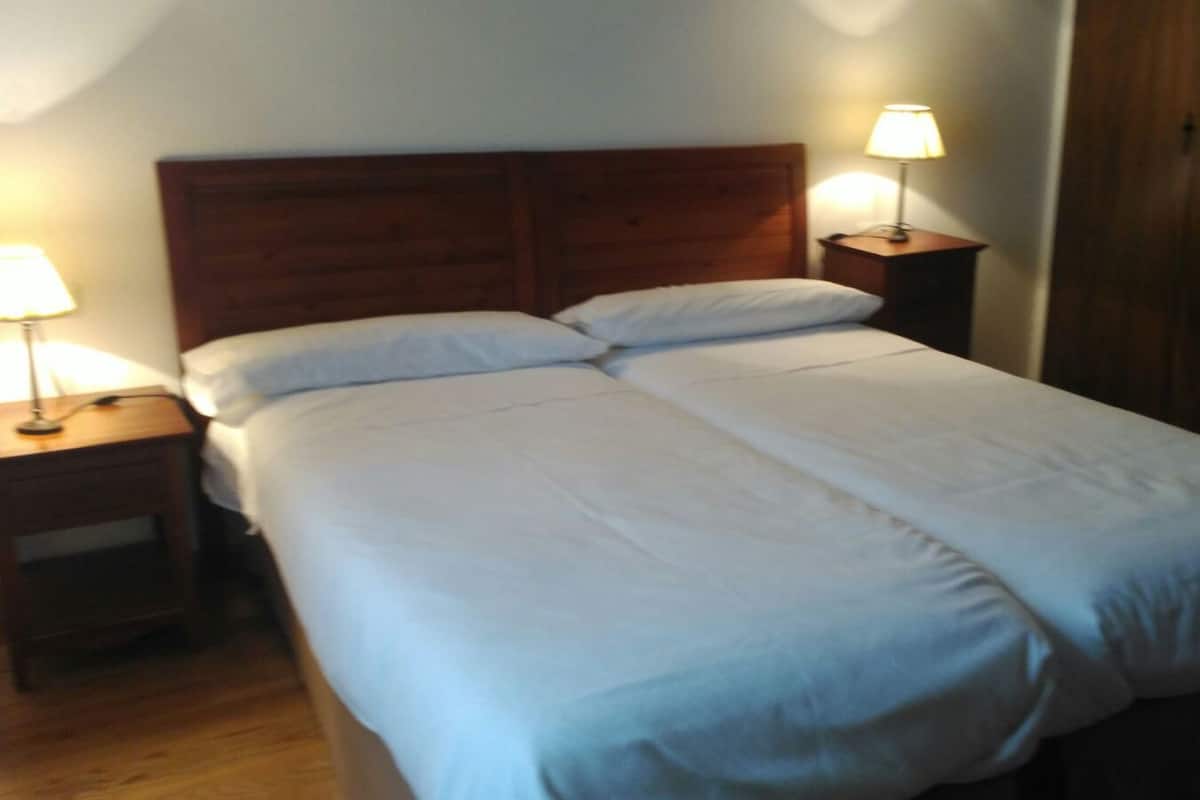 Uma cama de casal com roupa de cama branca, duas mesas de cabeceira com abajures e uma cabeceira de madeira.