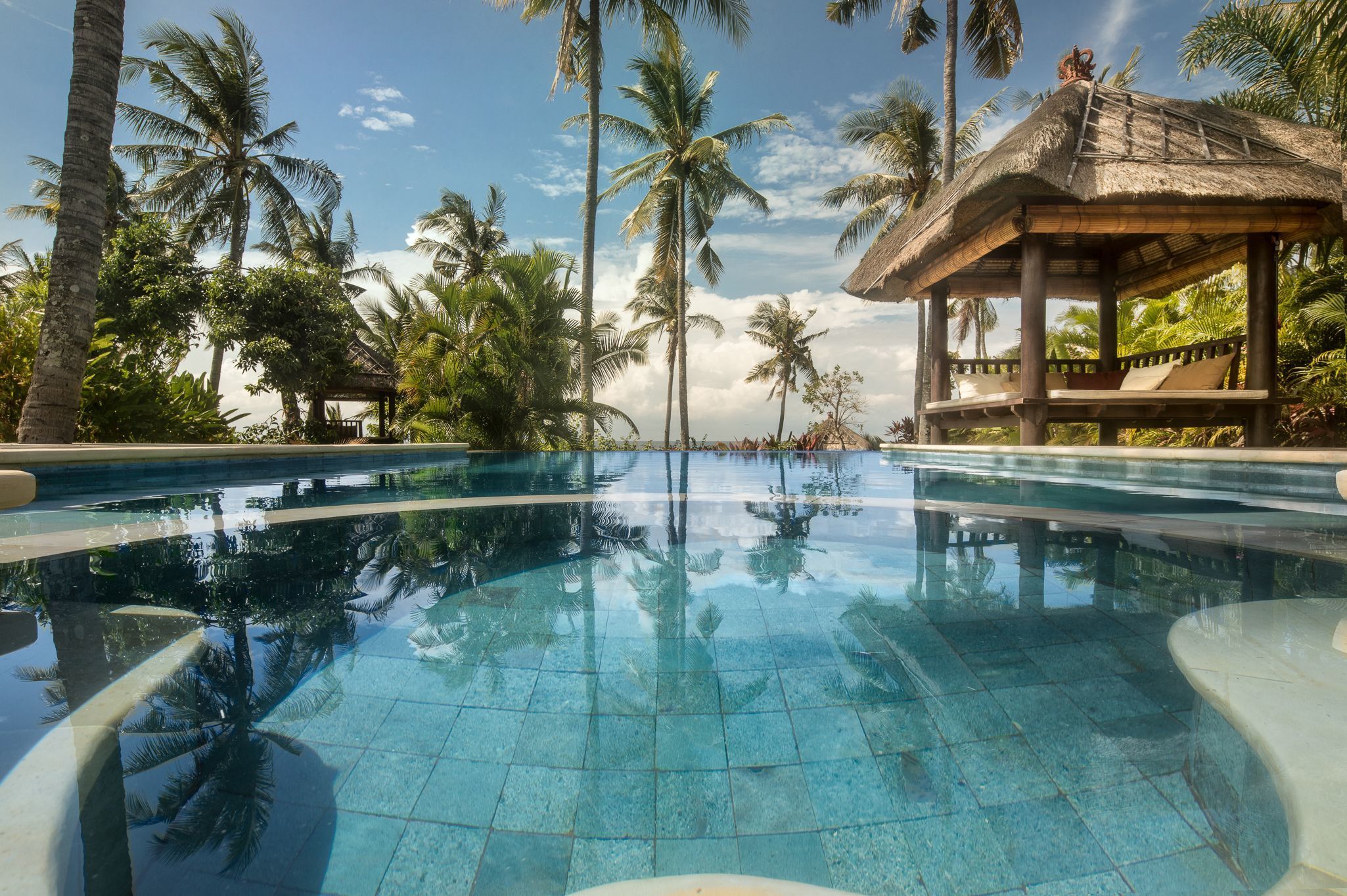 Uma piscina límpida refletindo palmeiras, um gazebo com telhado de palha e vegetação exuberante.