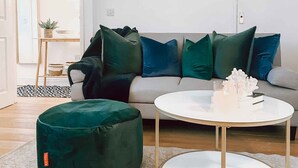 Uma sala de estar moderna com um sofá cinzento, almofadas verdes e azuis, uma mesa de centro branca redonda e um banquinho verde.