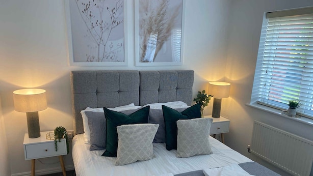 Uma cama bem arrumada com roupa de cama cinzenta e várias almofadas, duas mesas de cabeceira com abajures e uma janela com persianas.