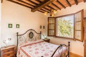 Quarto com teto de madeira, cama com colcha florida, mesa de cabeceira com abajur e janela com vista para árvores.
