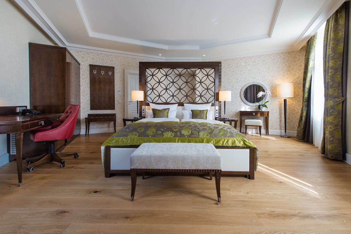Quarto de hotel com uma cama grande, uma escrivaninha com cadeira, uma luminária de mesa, mesa de cabeceira com abajur e um espelho.