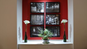 Uma janela com quatro fotografias a preto e branco numa parede vermelha, um vaso verde com flores brancas e duas garrafas verdes de cada lado.