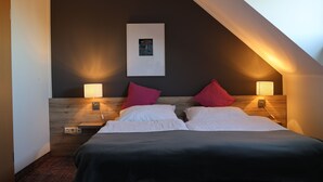Quarto de hotel com uma cama, duas luminárias laterais, um quadro na parede e um painel de cabeceira de madeira.
