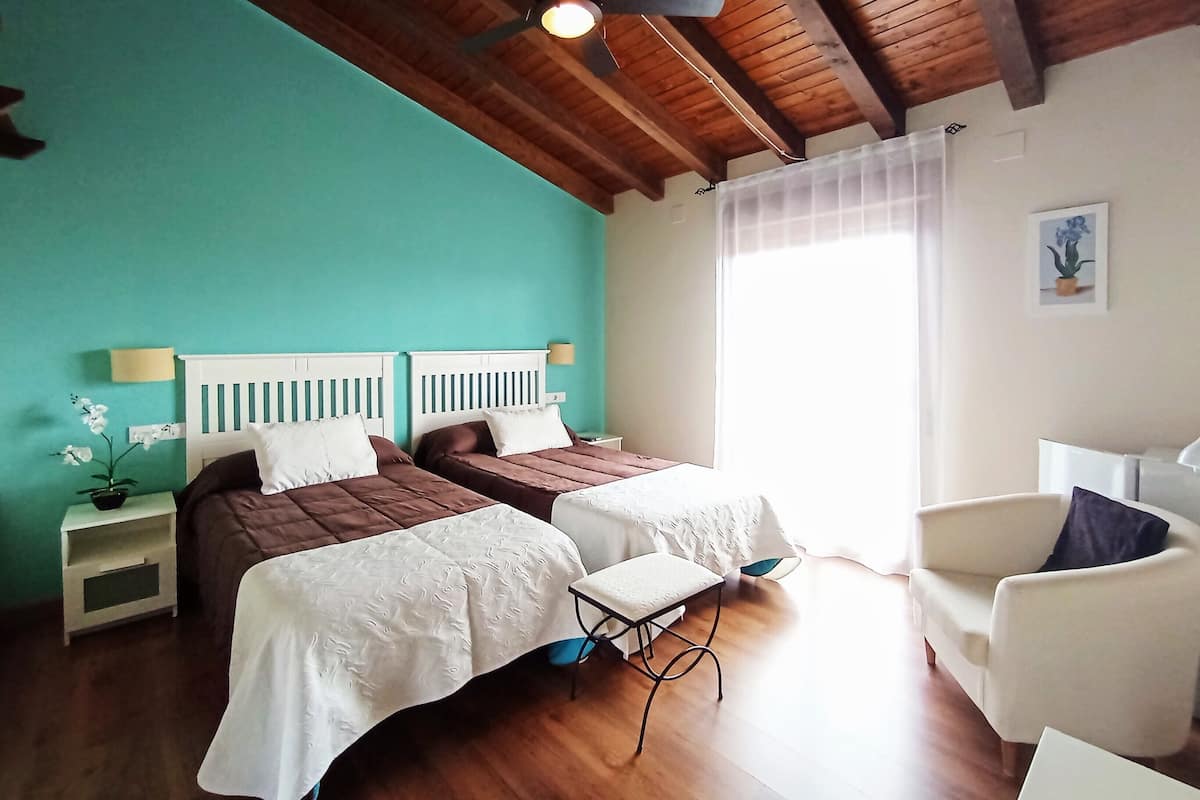 Quarto de hotel com duas camas, um ventilador de teto, uma janela com cortinas, uma cadeira branca e uma pequena mesa.