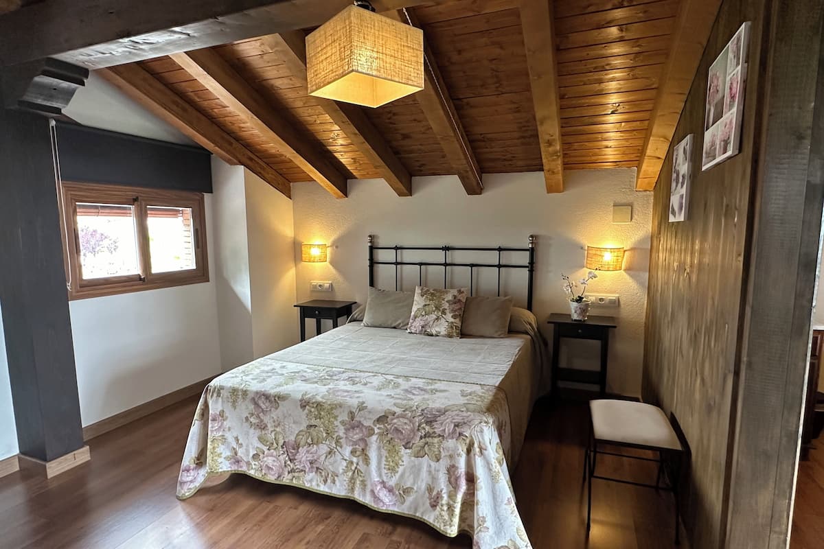 Quarto com teto de madeira, uma cama com roupa de cama floral, duas mesas de cabeceira com abajures e uma janela com persianas.