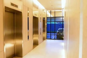 Um elevador moderno com visor digital, corredor bem iluminado com janela do chão ao teto e vista para um estacionamento.