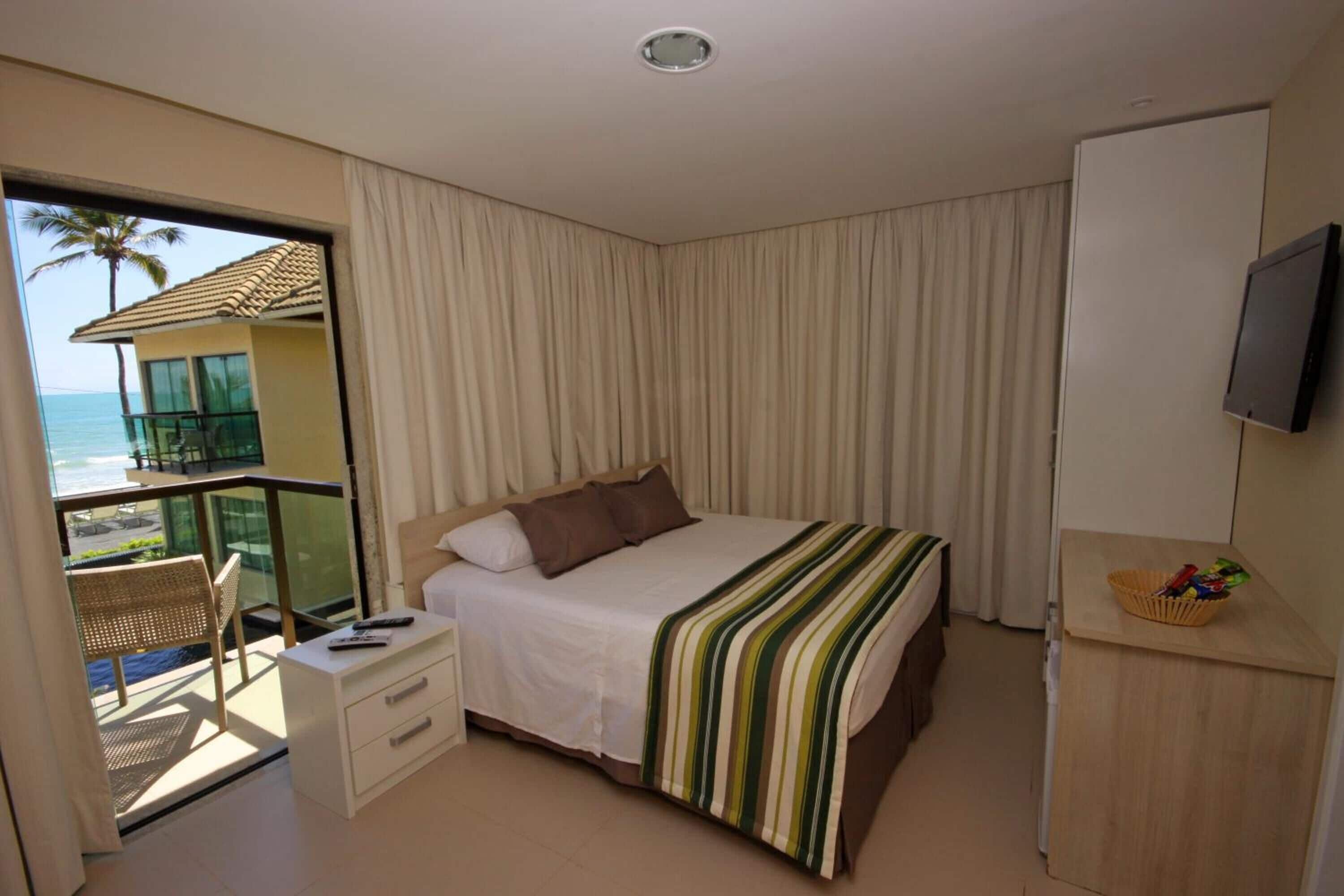 Quarto de hotel com cama, pequena escrivaninha, cadeira, televisão e varanda com vista para o mar e palmeiras.