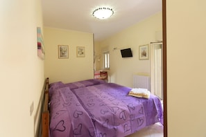 Quarto com cama coberta por um cobertor roxo com estampas de coração, mesa de cabeceira com luminária e janela com vista para um edifício.