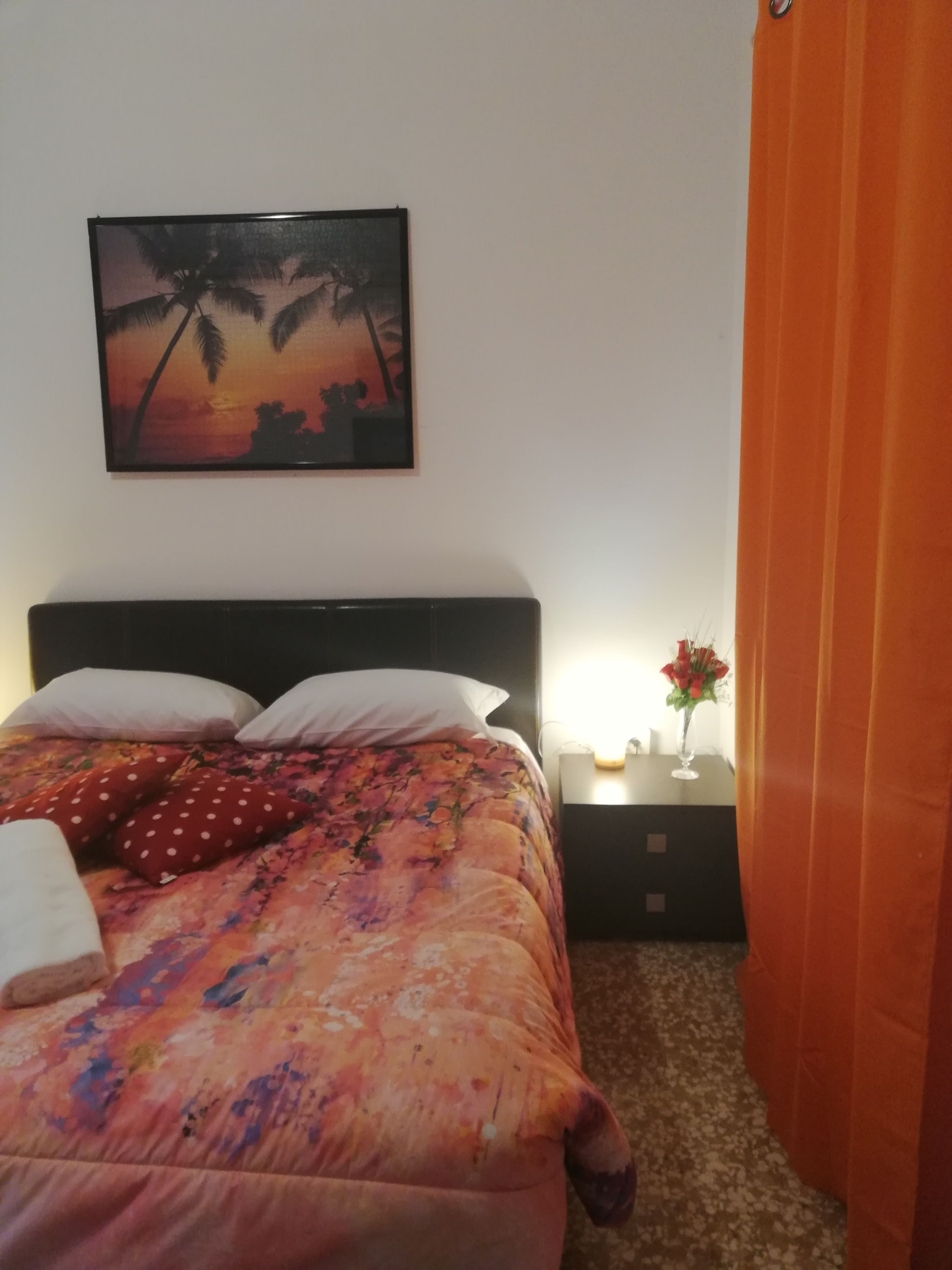 Uma cama com colcha florida, uma mesa de cabeceira com um vaso de flores e um quadro com palmeiras na parede.