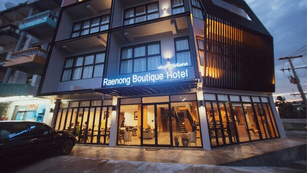 Um hotel moderno com uma placa indicando "Raenong Boutique Hotel". Apresenta uma estrutura em vários andares, com grandes janelas e uma escadaria visível.