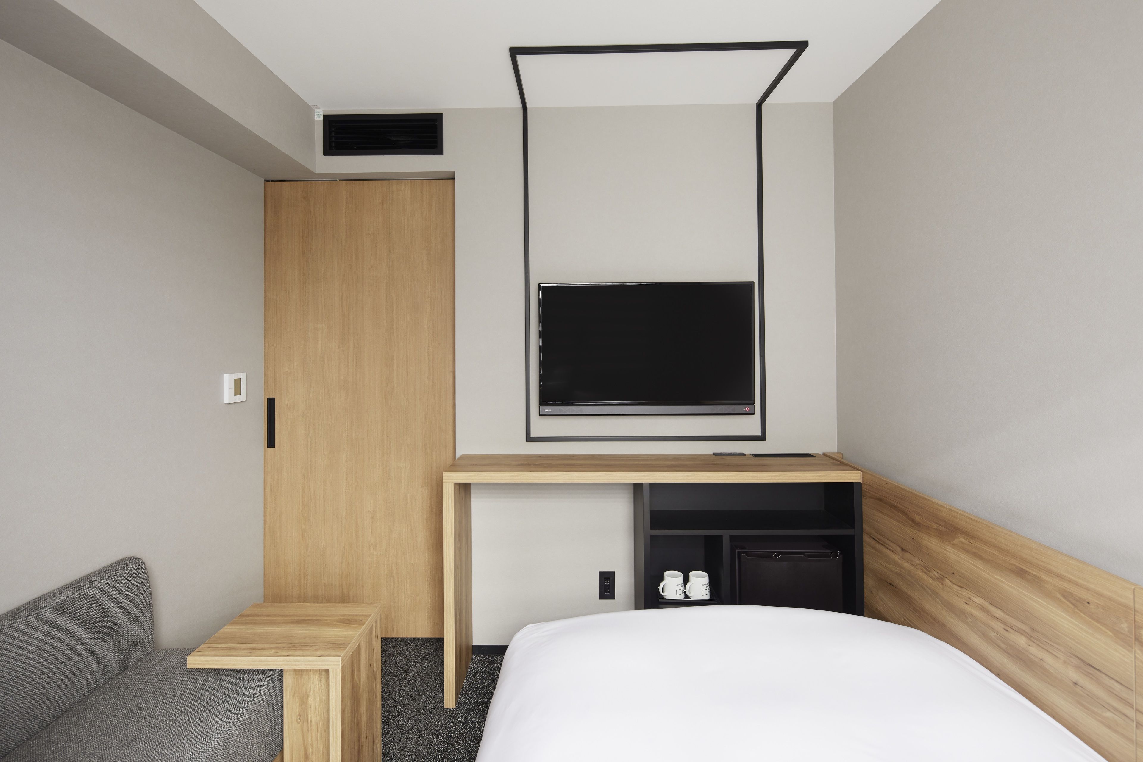 Quarto de hotel moderno com cama, sofá, escrivaninha com televisão de tela plana, guarda-roupa e uma pequena prateleira com xícaras.