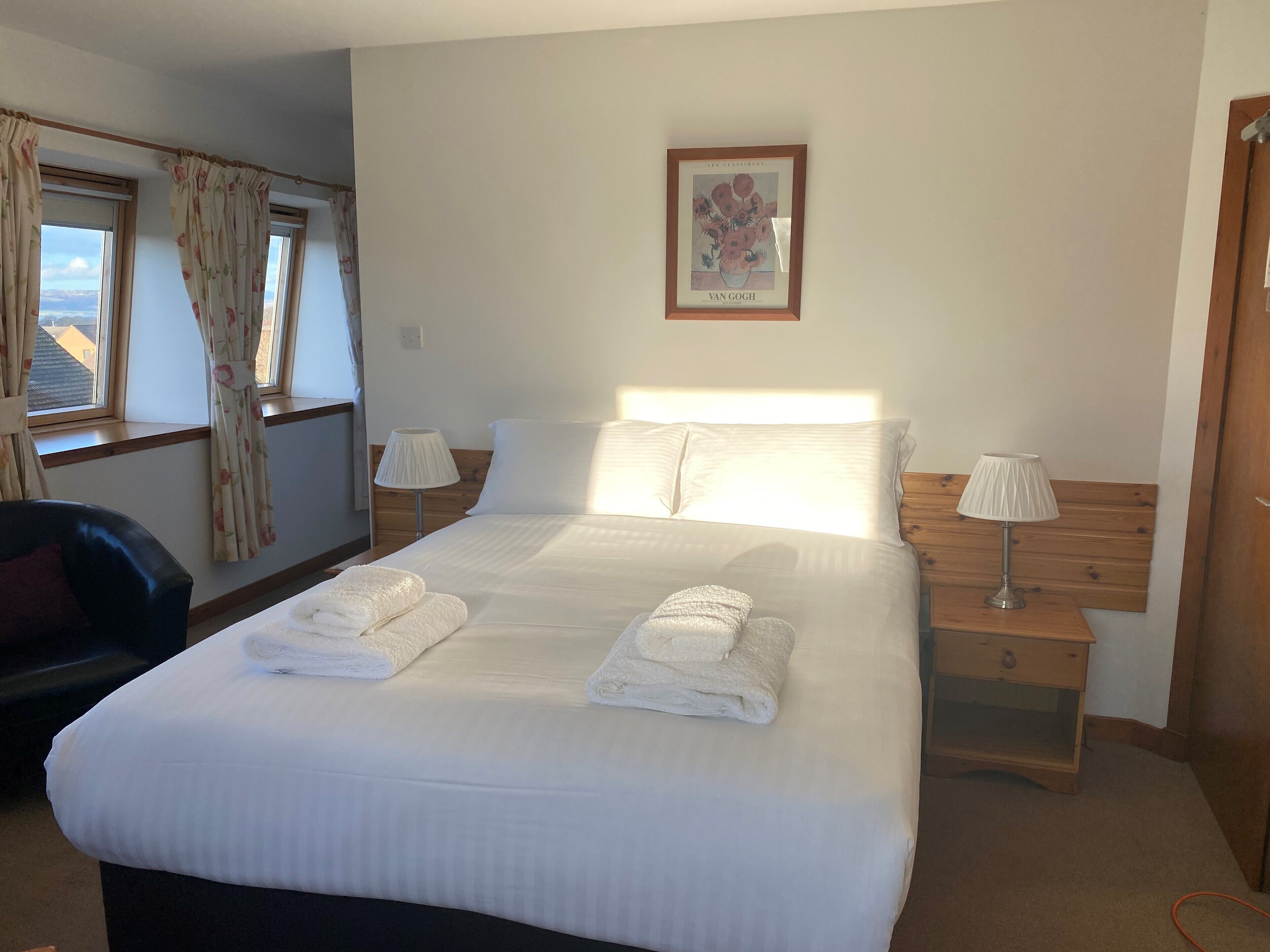 Cama bem arrumada com duas almofadas e duas toalhas dobradas, cabeceira de madeira, duas luminárias de apoio, quadro na parede e janela com cortinas.