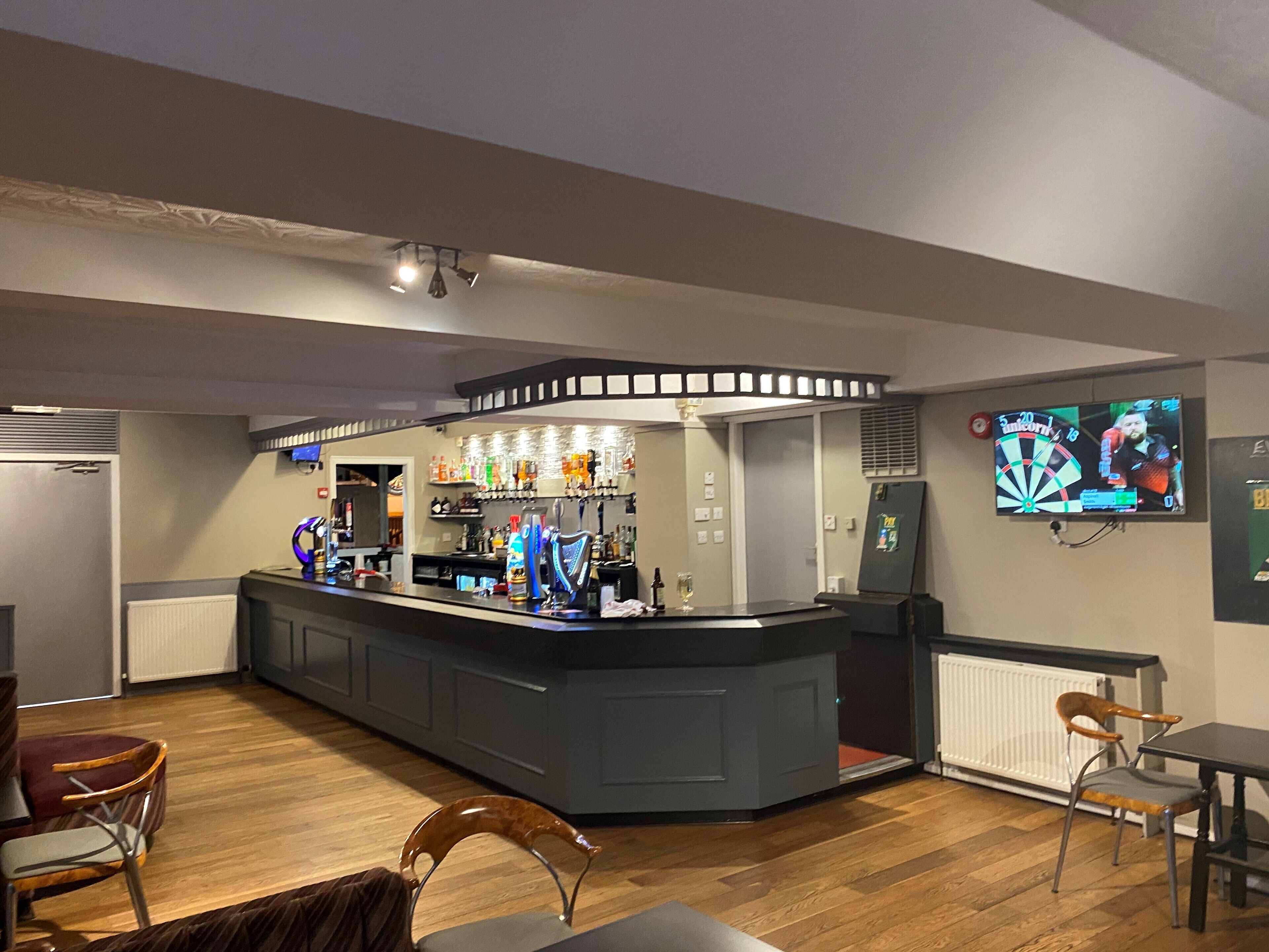 Uma área de bar com balcão, prateleiras com garrafas, alvo de dardos e uma tela de televisão.