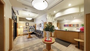 Uma área de recepção com balcão de check-in, um espaço de estar com cadeiras, um vaso decorativo com flores e piso revestido de azulejos.