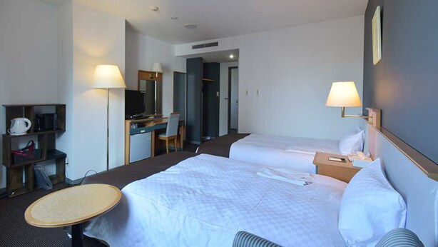 Quarto de hotel com duas camas, uma escrivaninha, uma cadeira, uma mesinha e um abajur.