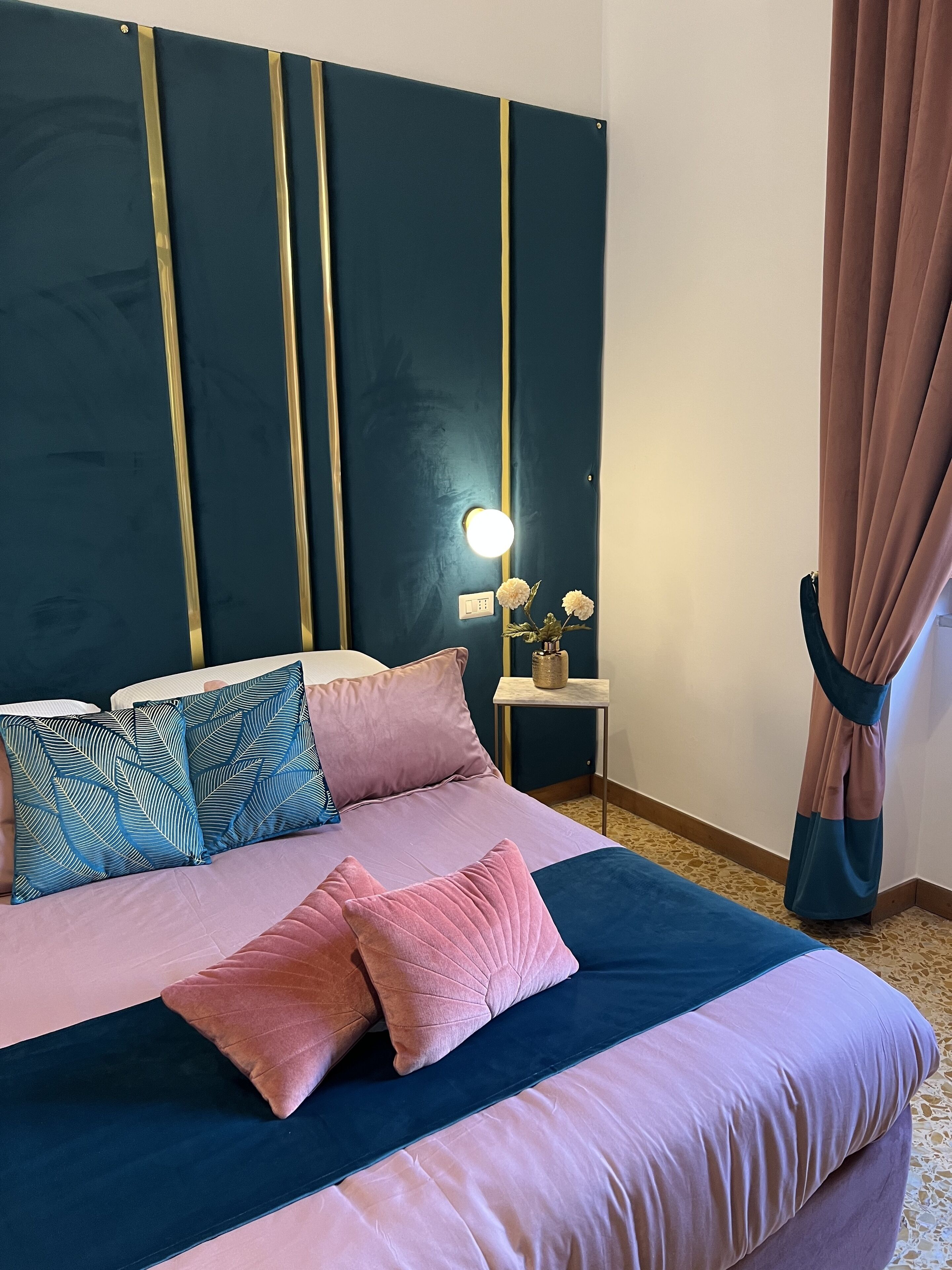Uma cama com colcha azul e rosa, várias almofadas, uma mesa de cabeceira com abajur e um vaso com flores.