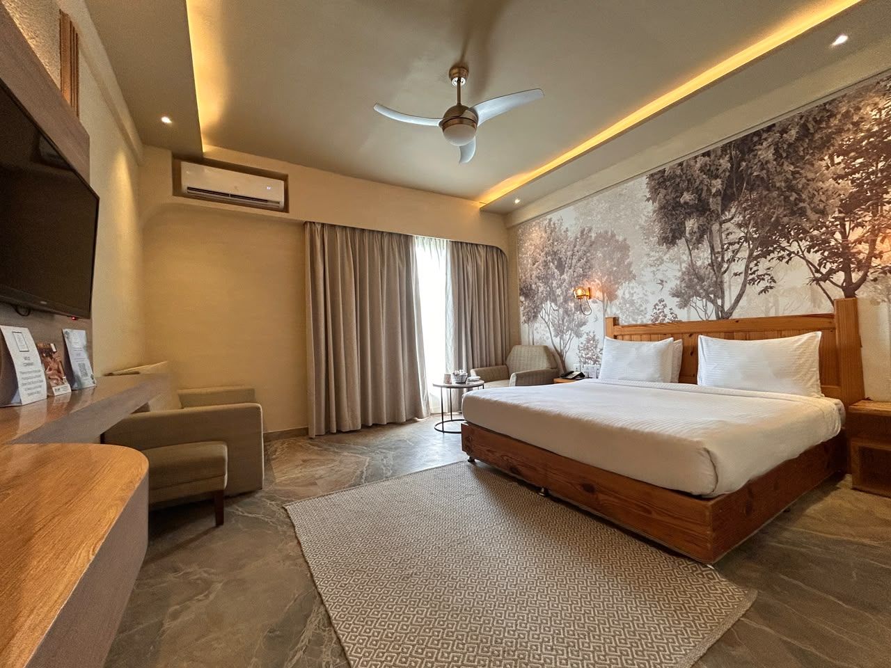 Quarto de hotel com cama, escrivaninha, cadeira, ventoinha no teto e ar condicionado fixado na parede.