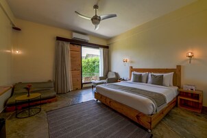 Quarto com uma cama grande, ventilador de teto, janela com cortinas, uma mesinha com um vaso e criado-mudo com abajur.