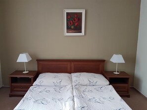 Quarto com cabeceira de madeira, duas mesas de cabeceira, duas luminárias brancas, colcha com padrão floral e um quadro na parede.