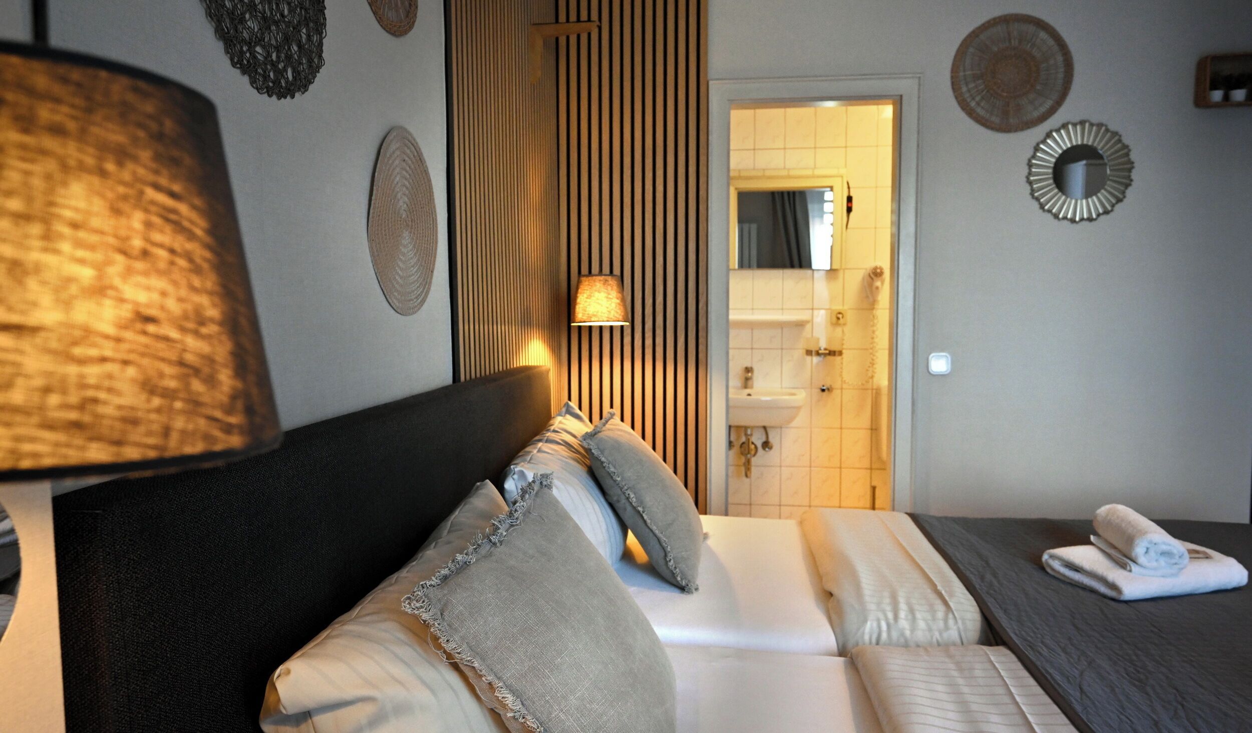 Quarto com cama, mesa de cabeceira com toalha, luminária e banheiro visível por uma porta.