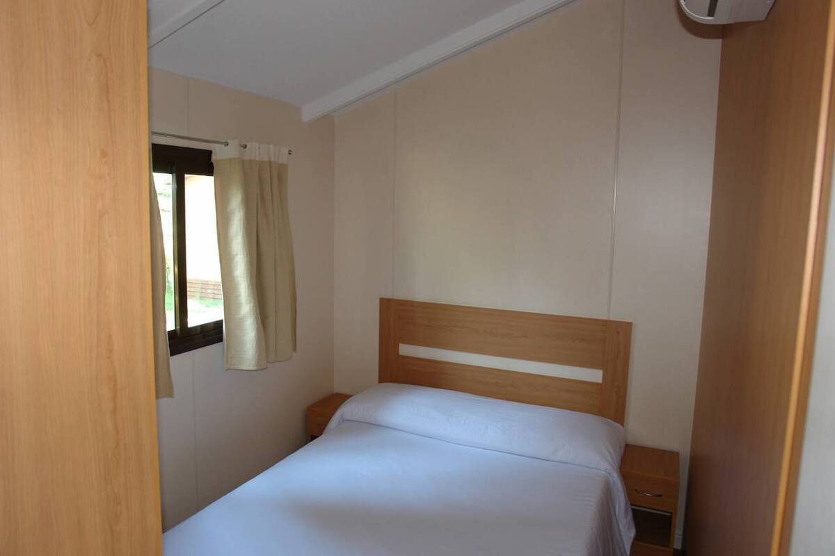 Cama de casal com cabeceira de madeira em quarto com paredes bege e janela com cortinas.