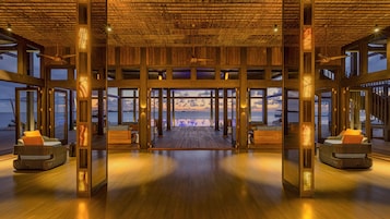 Uma sala de estar espaçosa e ao ar livre, com piso de madeira e áreas de estar, grandes janelas com vista para o mar e ventiladores de teto para ventilação.