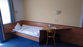 Quarto de hotel pequeno, com uma cama, uma escrivaninha e uma cadeira.