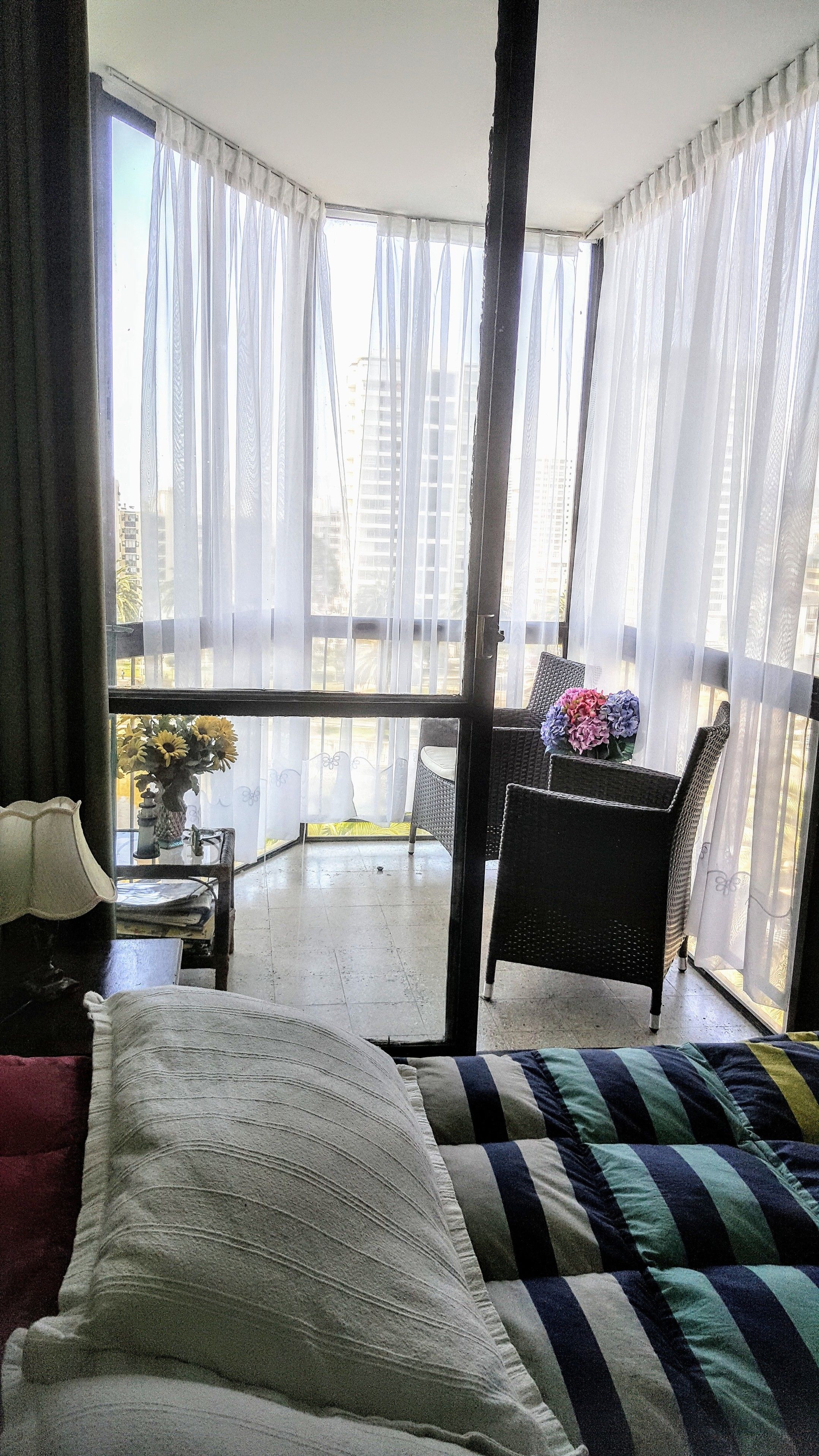 Quarto de hotel com uma cama grande, uma cadeira, uma mesa pequena com um vaso de flores e uma varanda com vista para edifícios.