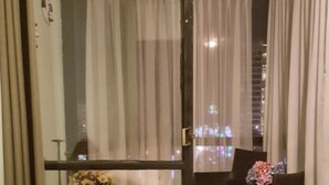 Quarto de hotel com uma cama, uma cadeira, uma mesa com um vaso de flores e vista para a cidade à noite através de uma porta de vidro.