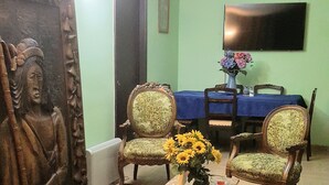 Uma sala de estar com duas cadeiras ornamentadas, uma mesa de centro com um vaso de girassóis, uma grande estátua de Buda, uma mesa de jantar com cadeiras e uma televisão de tela plana na parede.