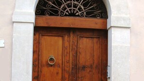 Uma porta dupla de madeira com um arco decorativo de metal acima, em fachada de um prédio de cor clara.