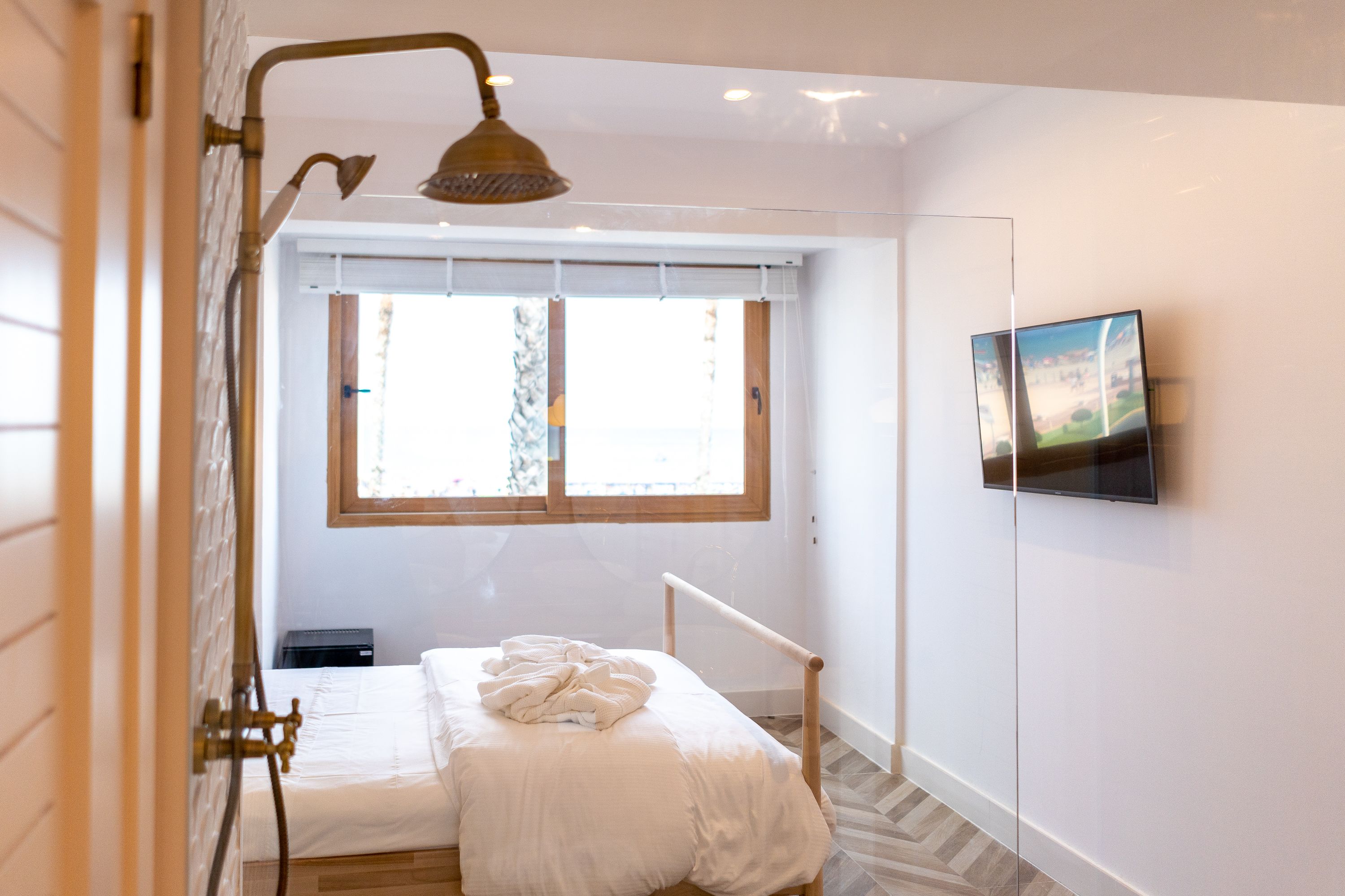 Quarto com cama, janela, TV fixada na parede e chuveiro com porta de vidro.
