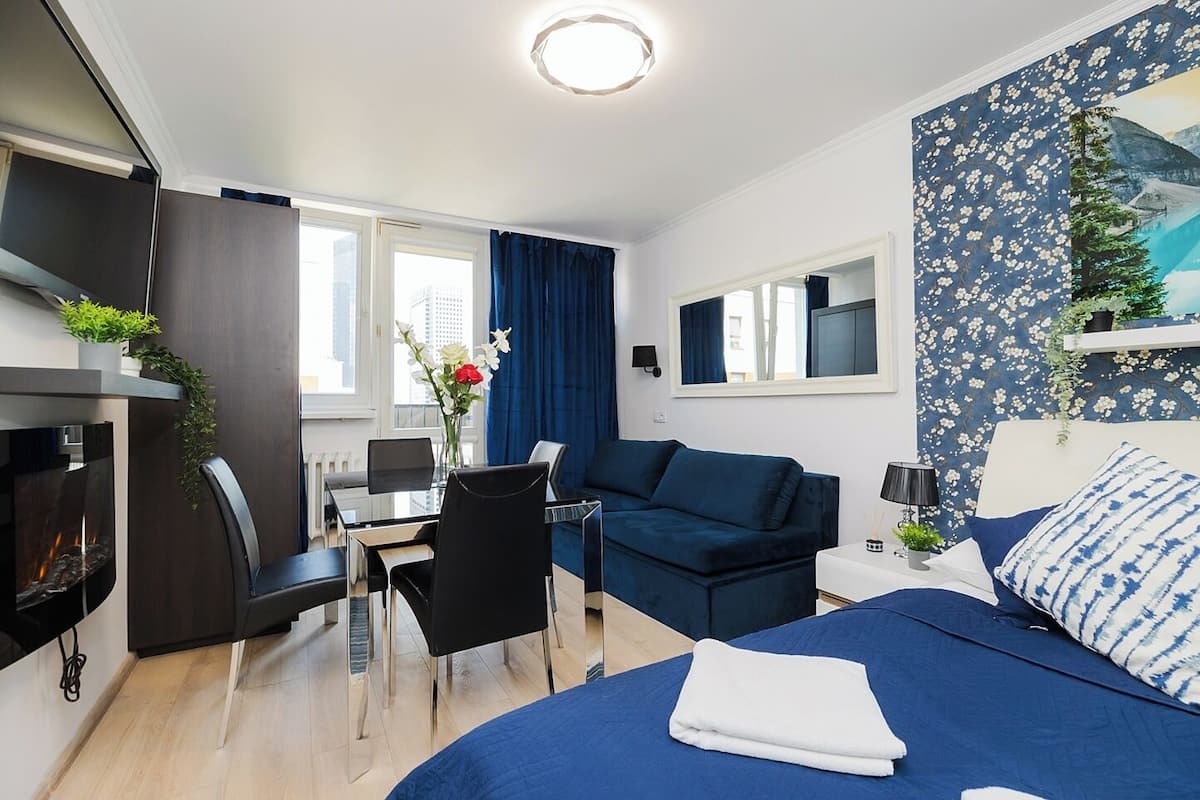 Quarto de hotel moderno com cama, sofá, escrivaninha com cadeiras, lareira e uma grande janela com cortinas.