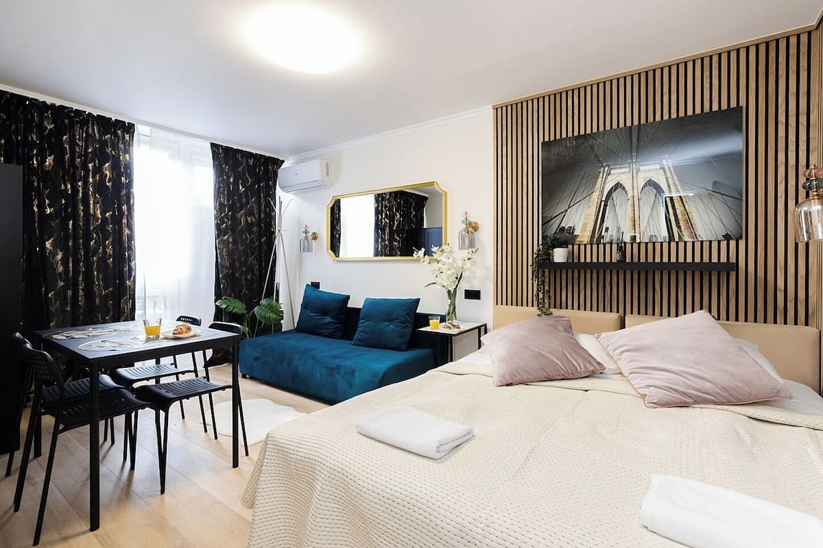 Quarto de hotel com cama, escrivaninha com cadeira, sofá, espelho e televisão fixada na parede.