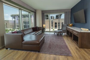 Quarto de hotel moderno com sofá de couro, uma escrivaninha, uma cadeira e grandes janelas com vista para a paisagem urbana.
