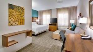 Quarto de hotel com cama, escrivaninha, cadeira, televisão e um quadro na parede.